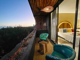 Varanda com vista para o mar, cama com roupa de cama branca e almofadas coloridas, e uma cadeira verde com padrão de estrelas.