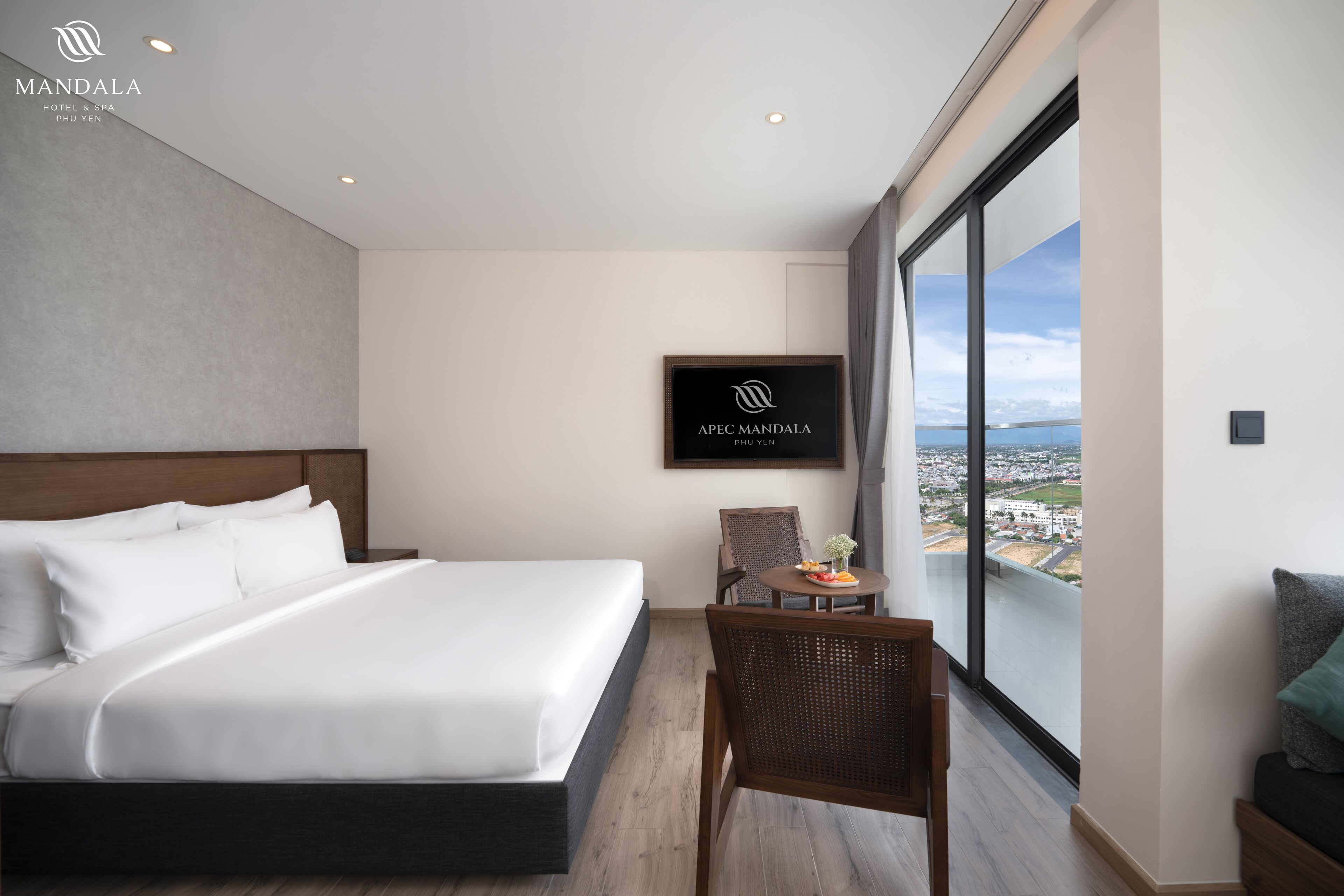 Quarto de hotel moderno com uma cama grande, uma mesinha com uma planta e vista para a cidade através de amplas janelas.