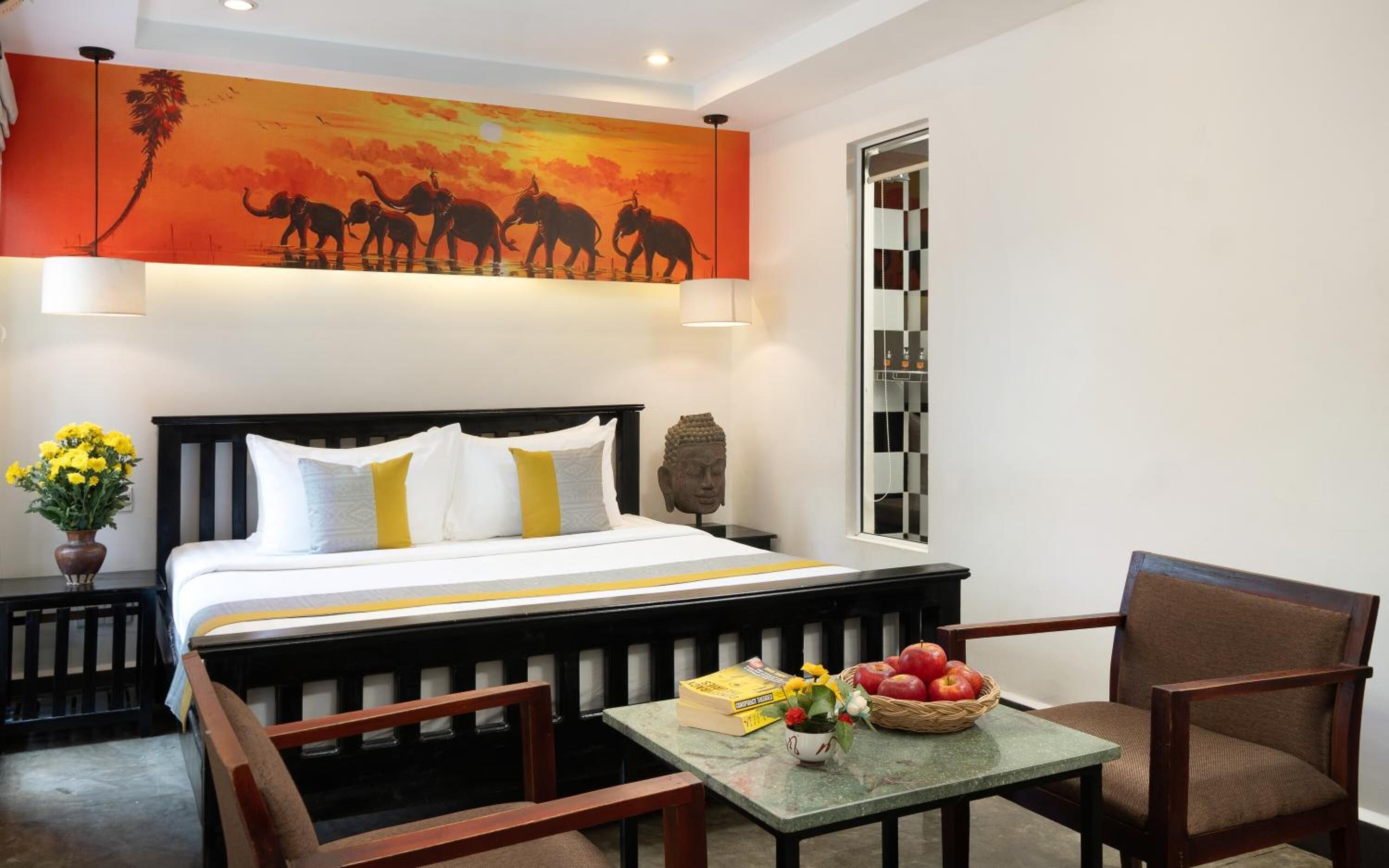 Quarto de hotel com cama, mesa de cabeceira, estátua de Buda, pintura de elefantes, um vaso de flores e uma mesa com maçãs e livros.