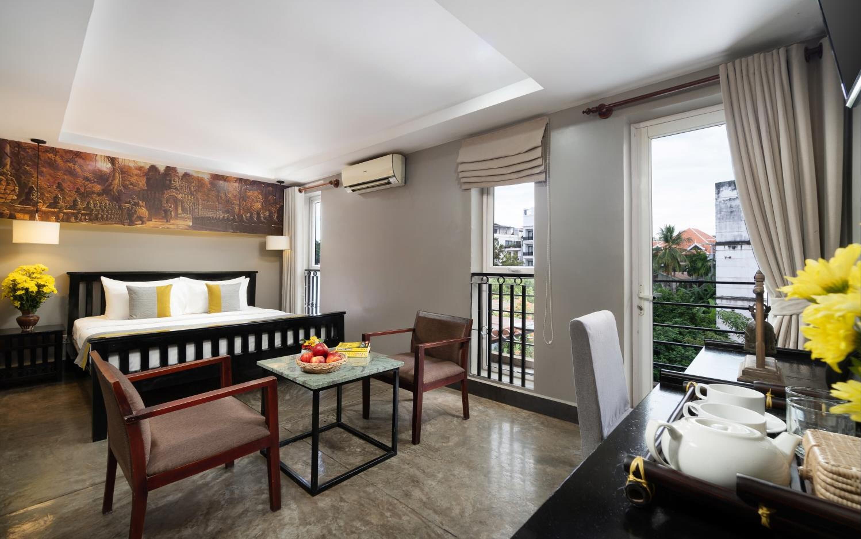 Quarto de hotel com cama, duas cadeiras, uma mesinha, varanda com grades e janela com cortinas.