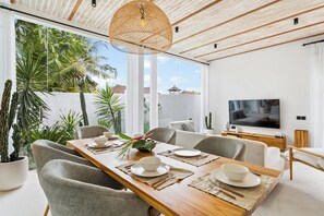 Uma sala de jantar moderna com mesa de madeira, cadeiras cinzas e uma grande janela com vista para vegetação e edifícios.