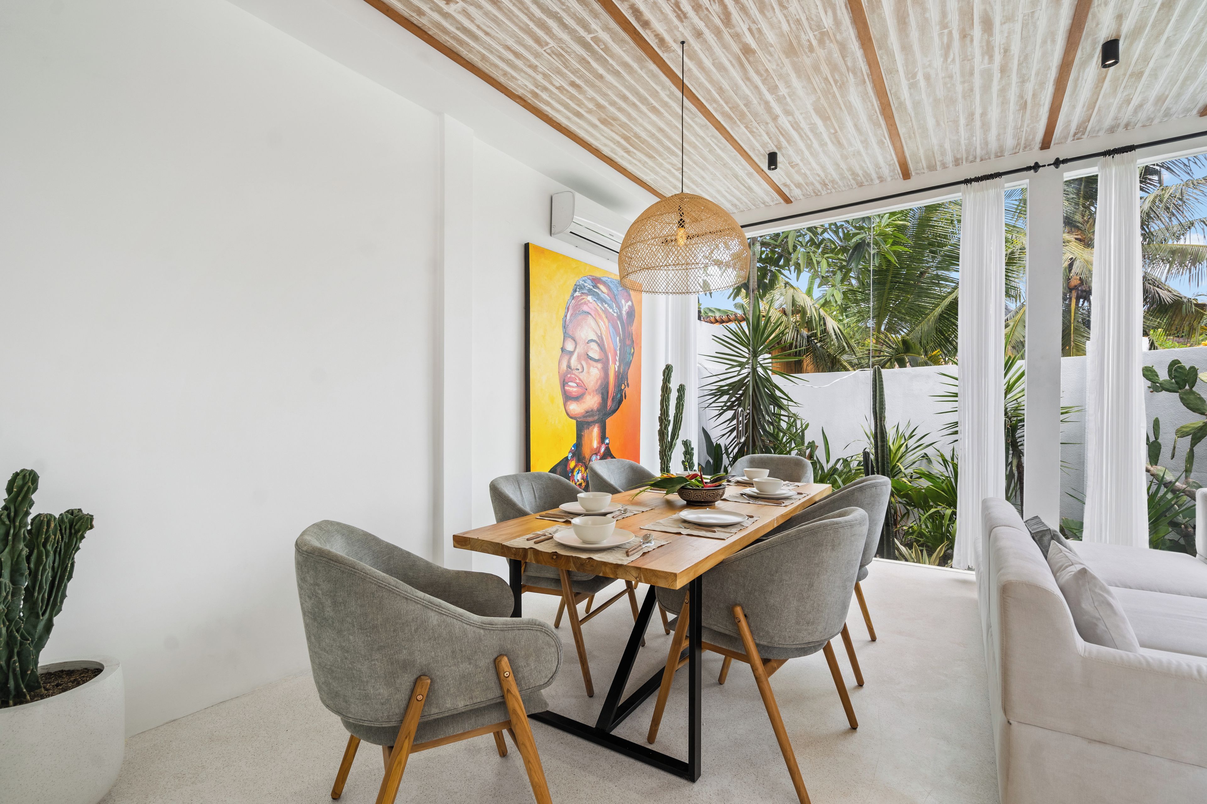 Uma sala de jantar com mesa de madeira, cadeiras estofadas cinzas e uma janela grande com vista para o verde.