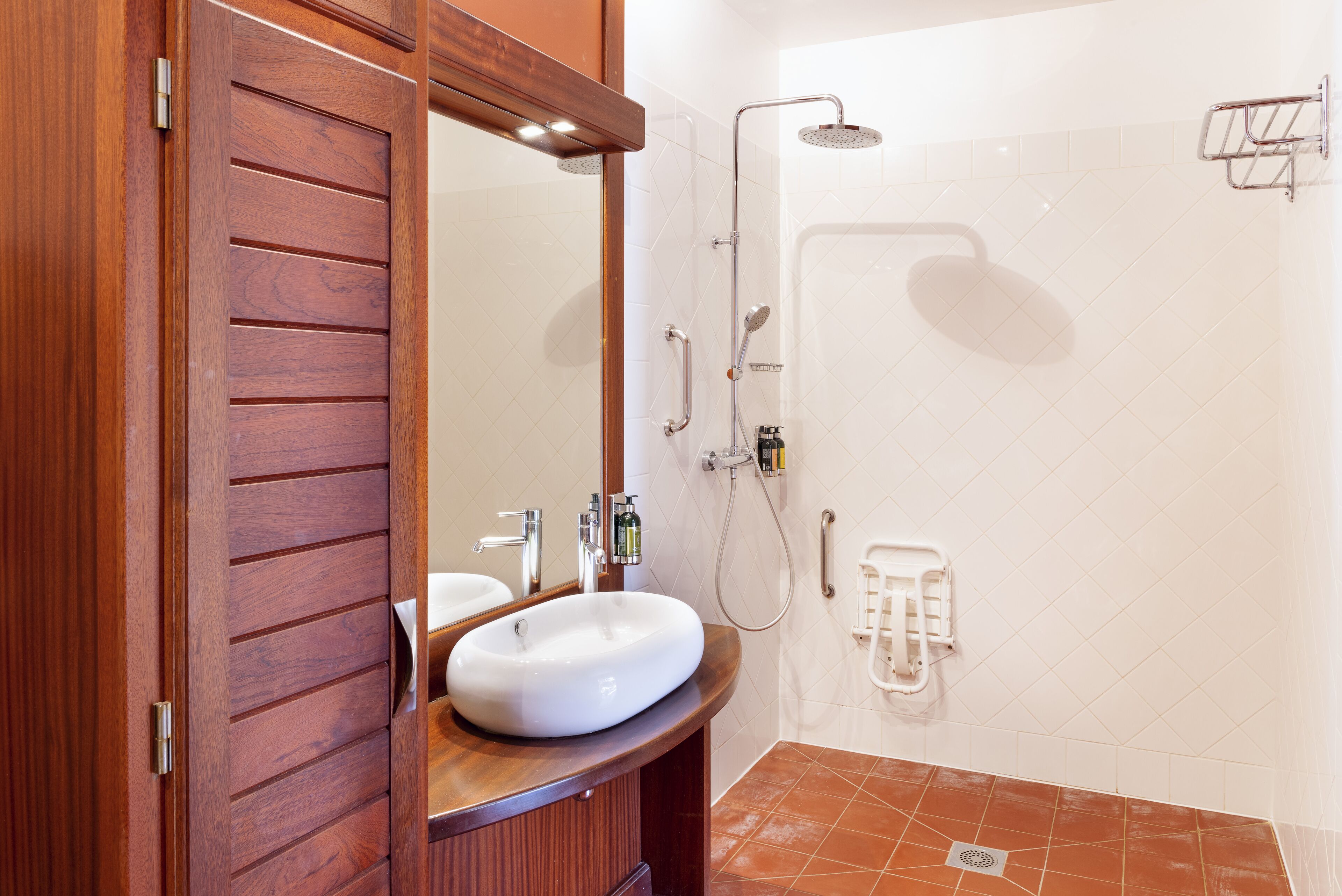Casa de banho com toucador de madeira, lavatório branco, espelho e zona de duche com chuveiro manual.