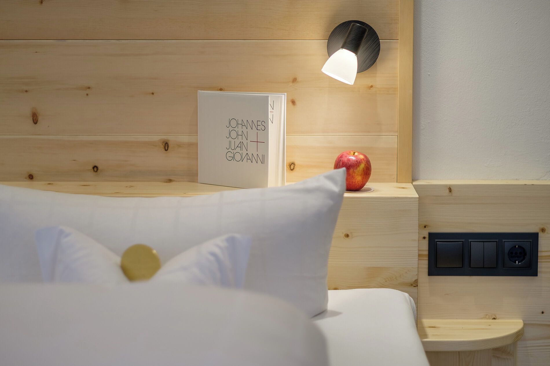 Cama bem arrumada com lençóis brancos e almofada com detalhe amarelo. Uma pequena mesa de cabeceira de madeira com uma placa branca e uma maçã vermelha são visíveis.