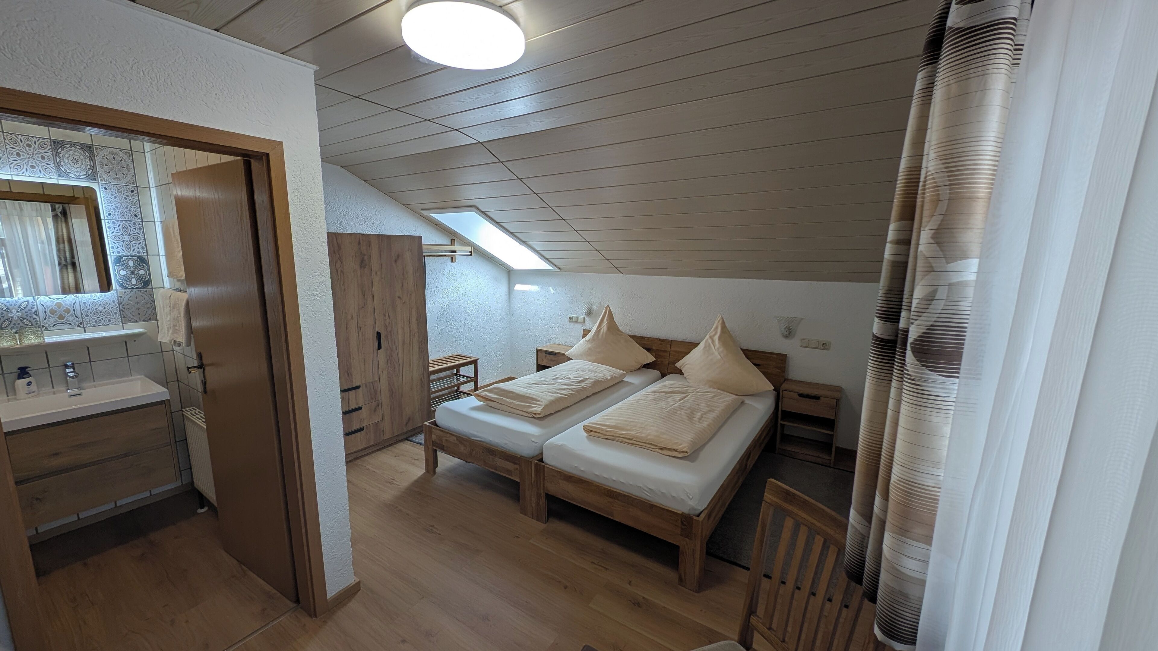 Quarto com duas camas, guarda-roupa de madeira, banheiro com pia e espelho, e um claraboia.