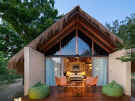 Uma construção moderna com telhado de palha e uma grande porta de vidro que dá acesso a um quarto com cama, mesas de cabeceira e luminária pendente.