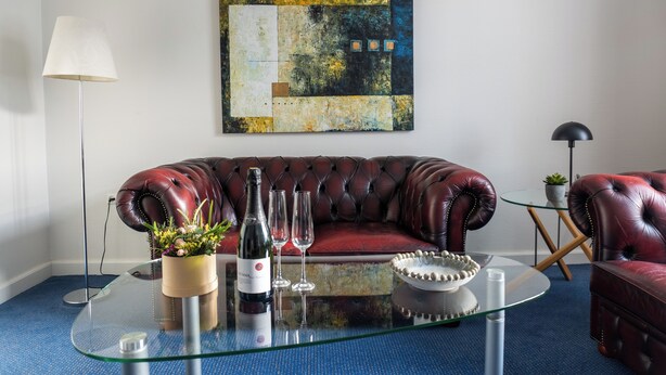 Uma sala de estar com um sofá de couro estofado, uma mesa de centro de vidro, um abajur e um quadro na parede.