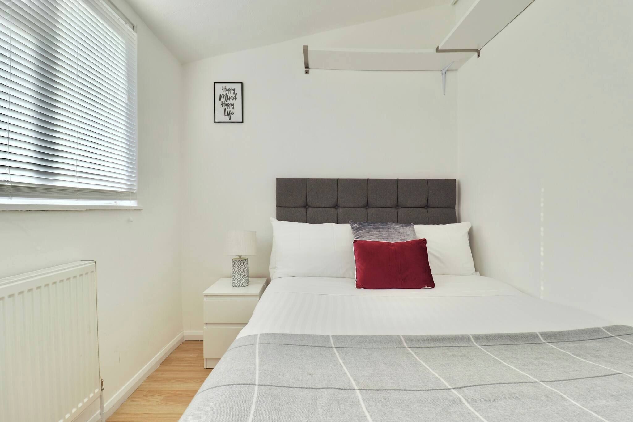 Quarto com cama, paredes brancas, radiador, mesa de cabeceira, abajur e janela com persianas.