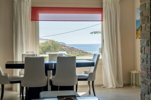 Uma área de refeições com mesa e cadeiras, vista para o mar e colinas, e uma parede de pedra.
