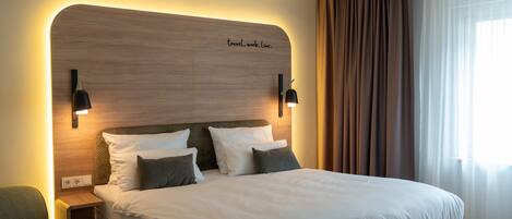 Quarto de hotel moderno com uma cama grande, duas mesinhas de cabeceira, uma cabeceira com a frase "travel. work. live.", e uma janela com cortinas.