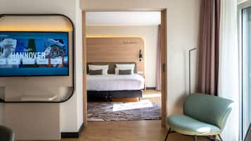 Quarto de hotel com cama, televisão na parede, uma cadeira e cabeceira de madeira.