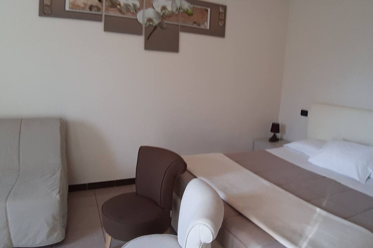 Quarto de hotel com cama, uma cadeira, mesa de cabeceira e um quadro na parede.