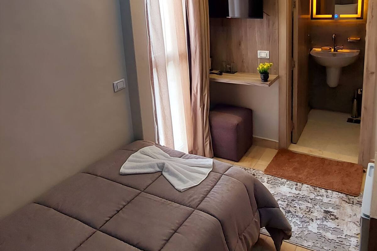 Quarto moderno com uma cama, uma penteadeira com espelho, e um banheiro visível através de uma porta aberta.