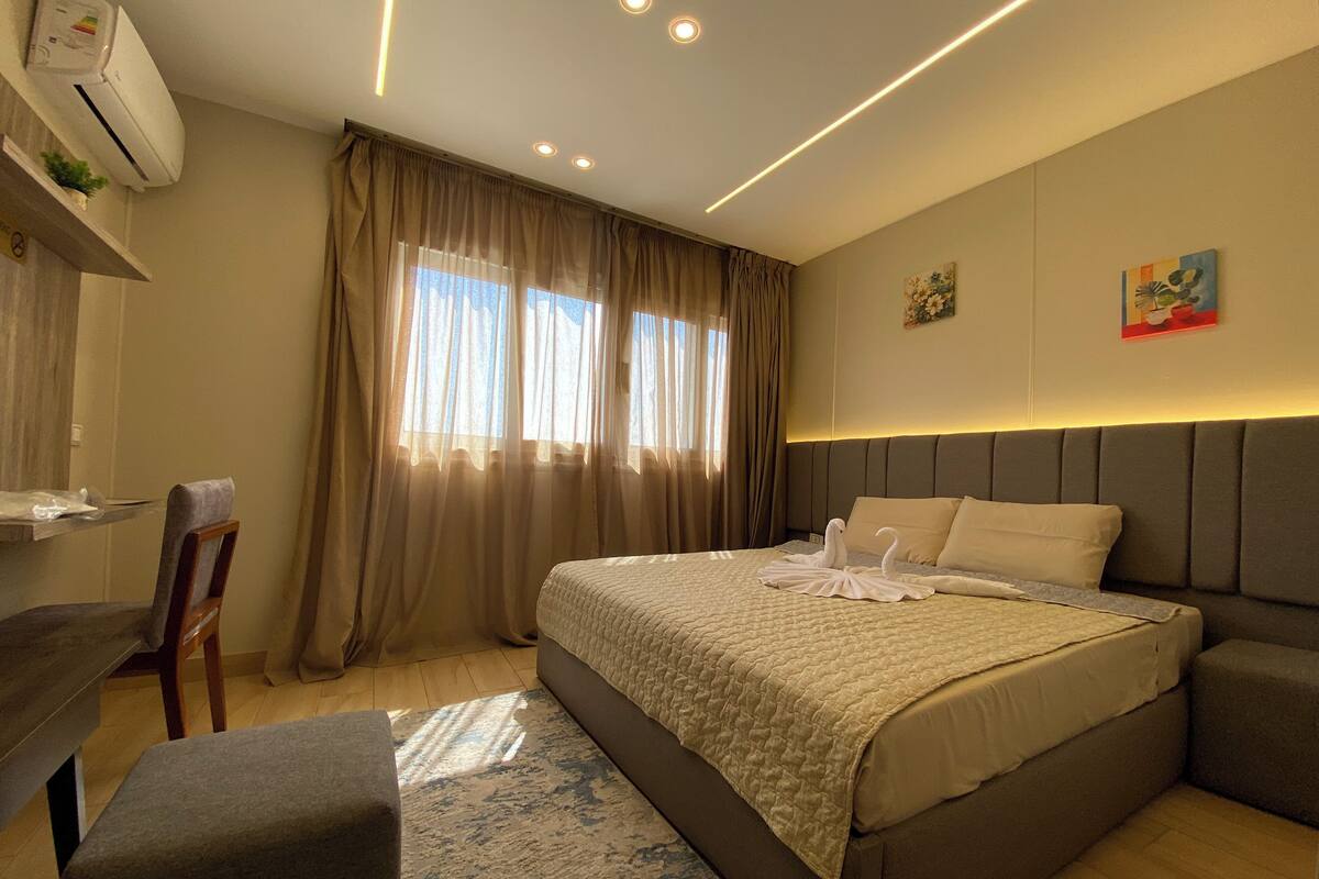 Quarto de hotel com uma cama, uma escrivaninha e uma cadeira. Há cortinas, uma janela e uma parede com um quadro.