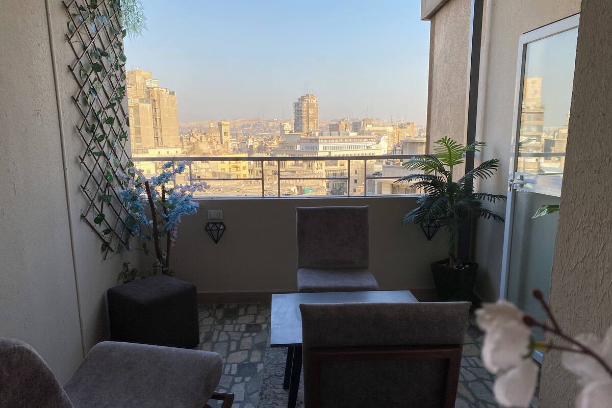 Varanda com vista para a paisagem urbana, apresentando uma mesa, duas cadeiras, um vaso de planta e um painel decorativo na parede.