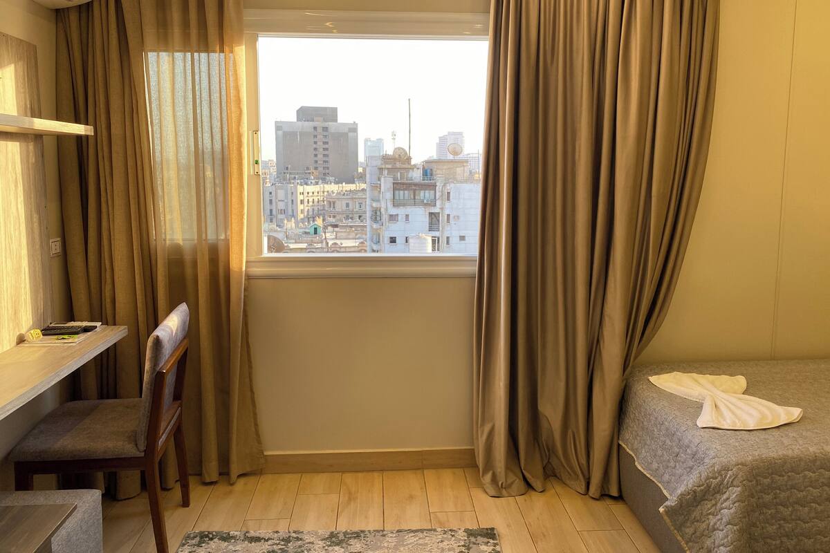 Quarto de hotel com uma janela grande, cama, escrivaninha e cadeira.