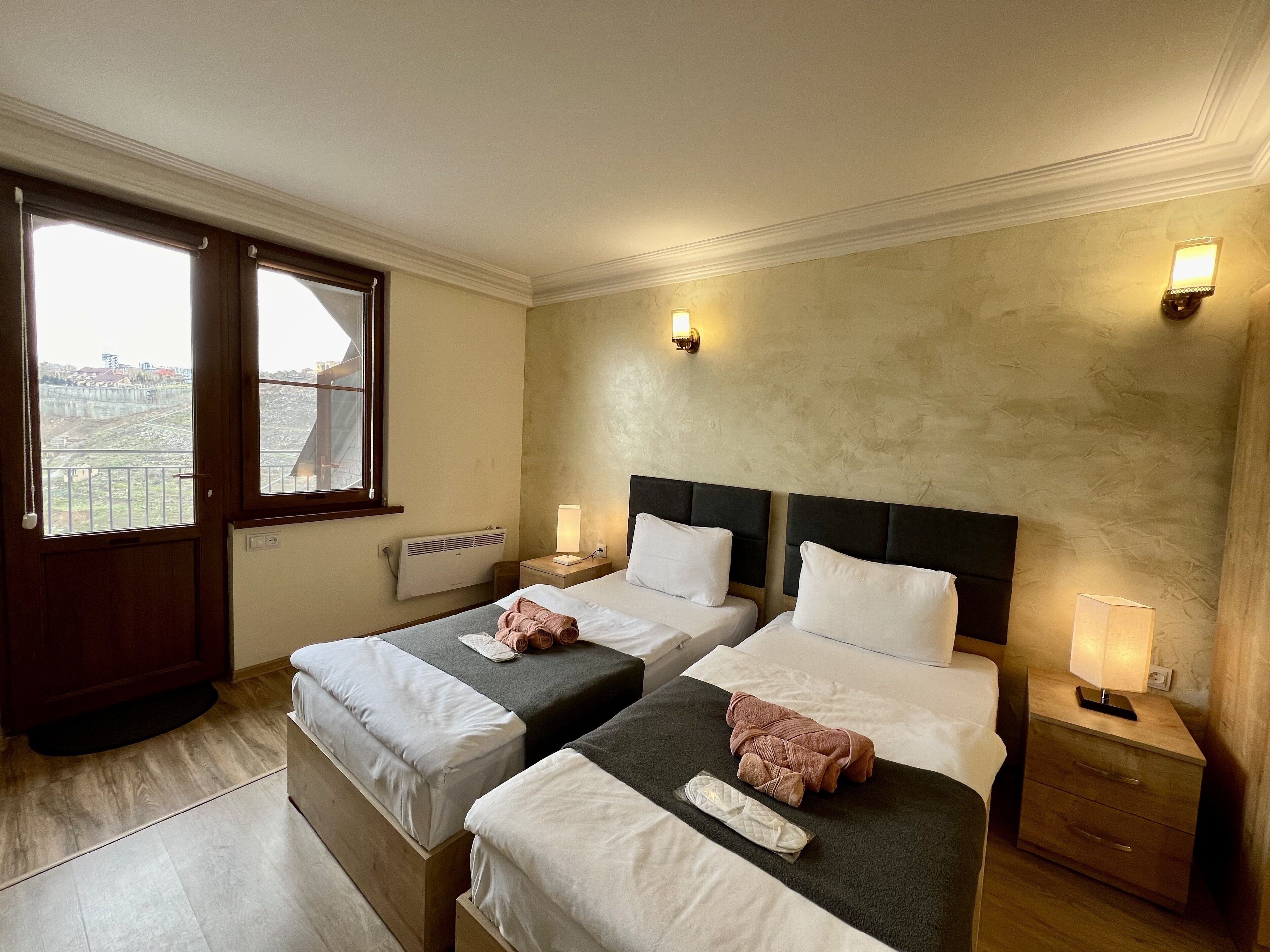Quarto de hotel com duas camas, uma janela, um radiador e uma mesa de cabeceira com abajur.