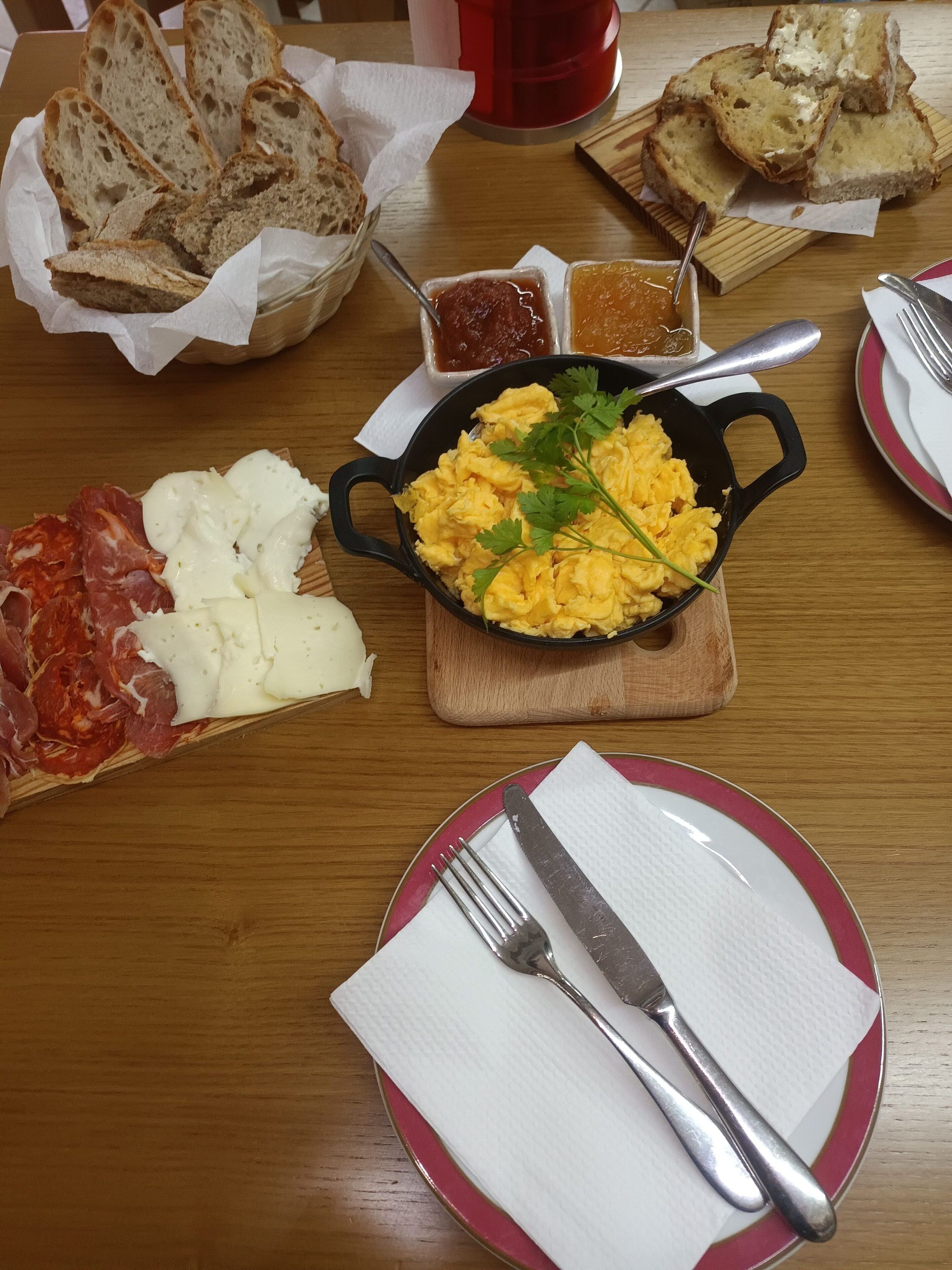Uma mesa de café da manhã com pão, ovos mexidos, queijo e embutidos.