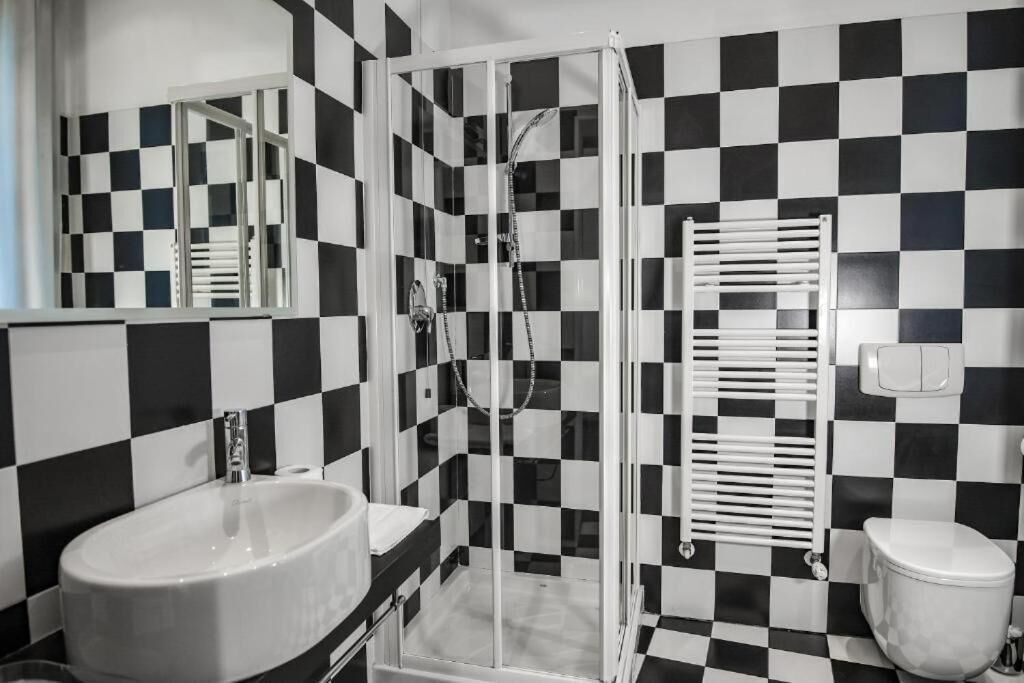 Casa de banho com padrão de azulejos preto e branco em xadrez, uma pia branca, chuveiro com parede de vidro e sanita branca.