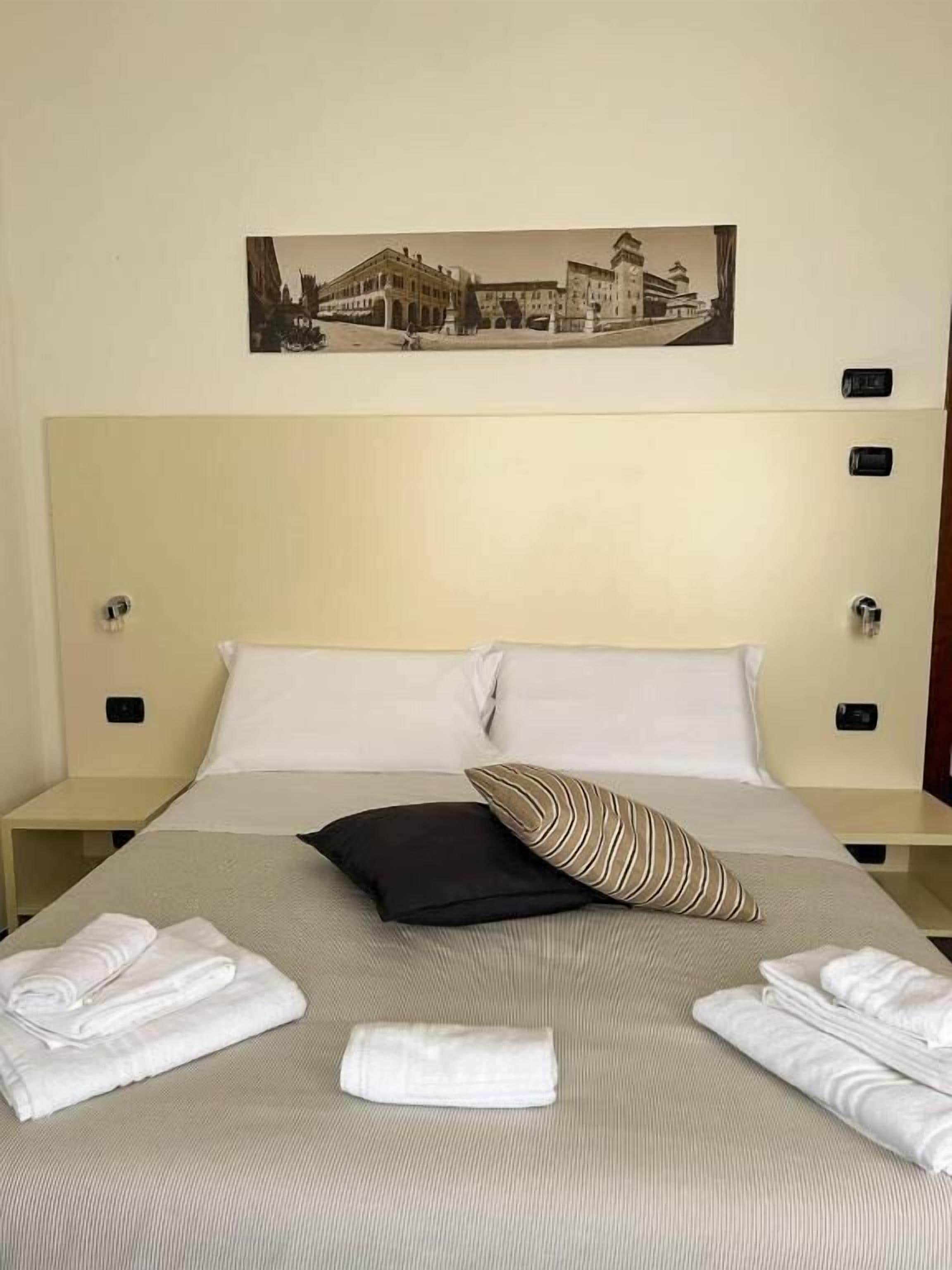 Cama bem arrumada com lençóis brancos e almofada listrada, mesa de cabeceira pequena com abajur, e quadro pendurado acima da cama.