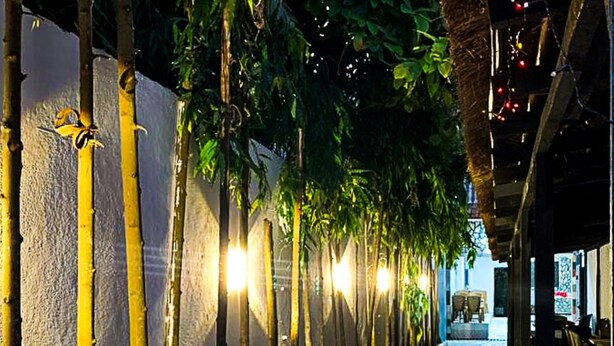 Uma passagem estreita ladeada por bambus verticais e iluminada por luzes quentes, conduzindo a um edifício com entrada visível.