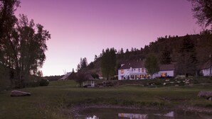 Uma casa grande e branca com varanda e um pequeno gazebo, cercada por árvores e um lago.