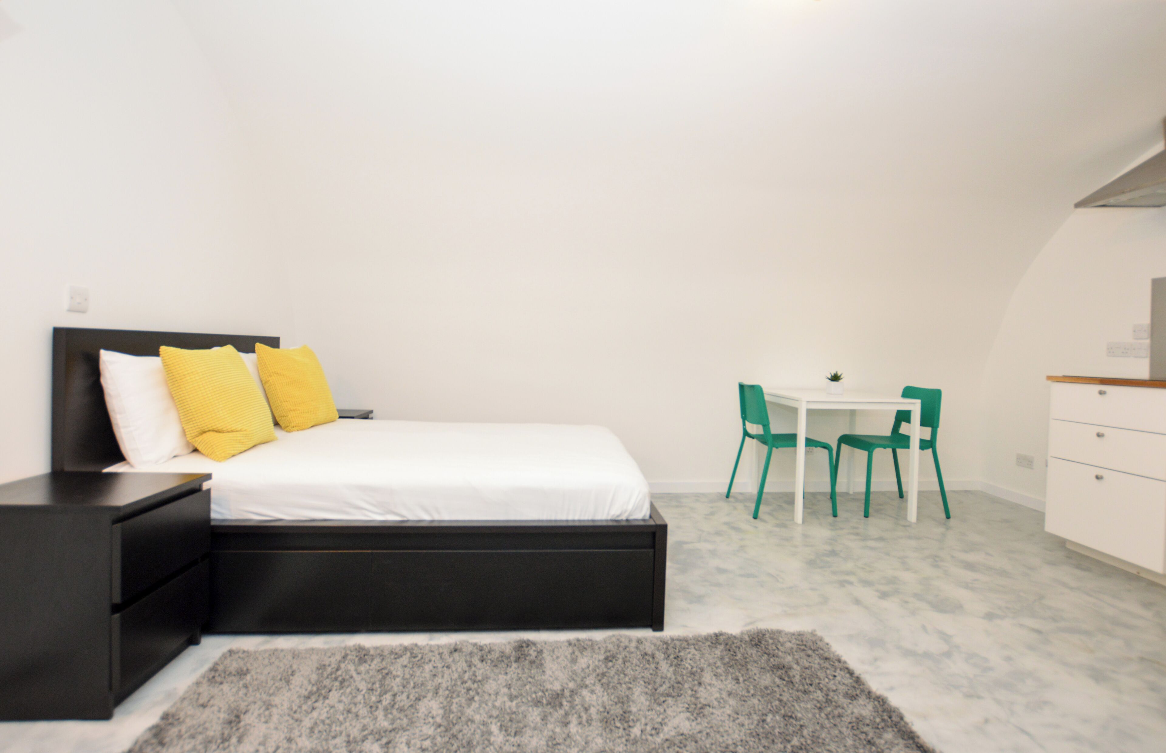 Um quarto com cama, mesa de cabeceira, escrivaninha com duas cadeiras e cômoda.
