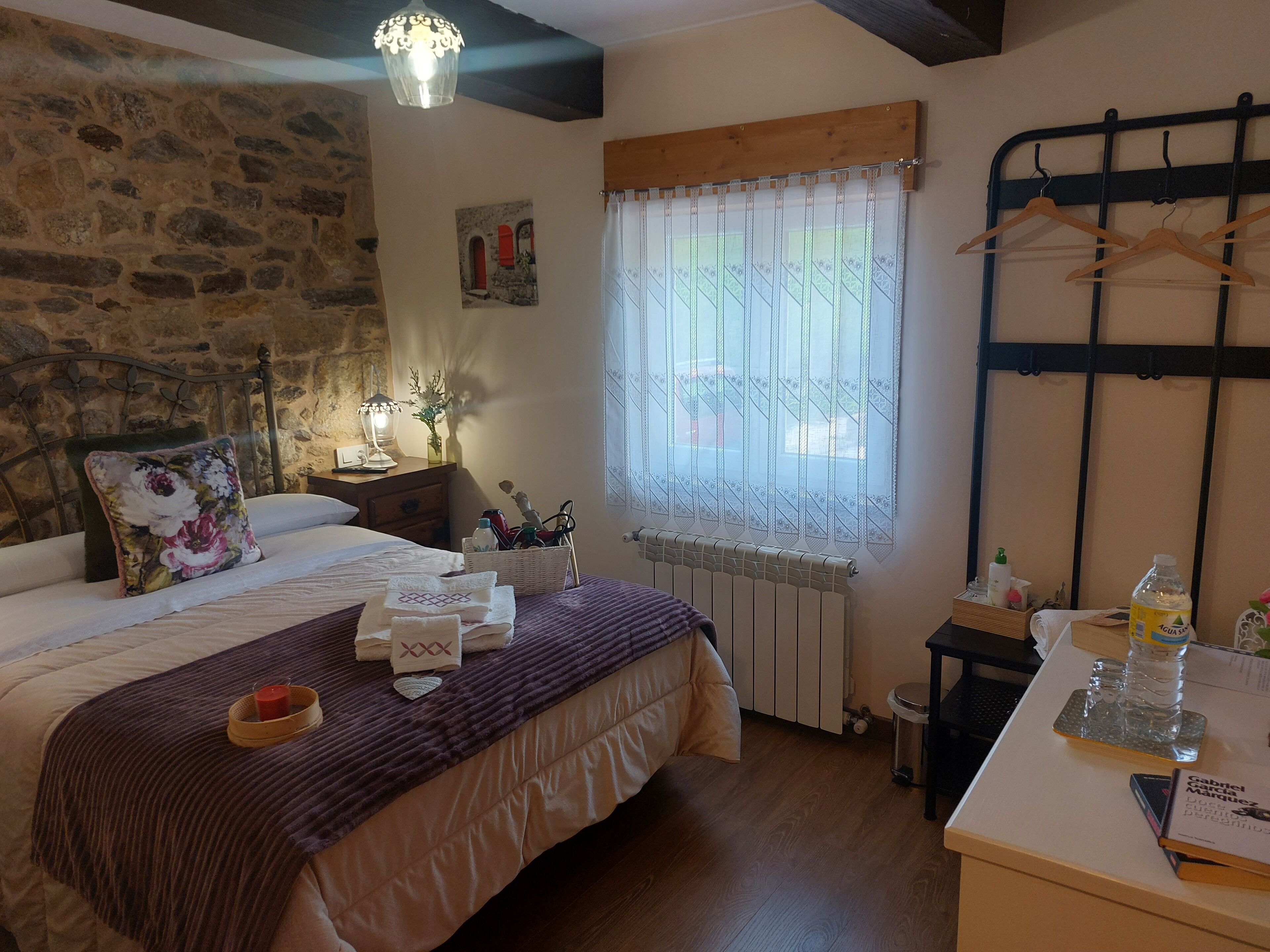 Um quarto aconchegante com parede de pedra, uma cama com travesseiro floral, mesa de cabeceira com abajur e janela com cortinas leves.
