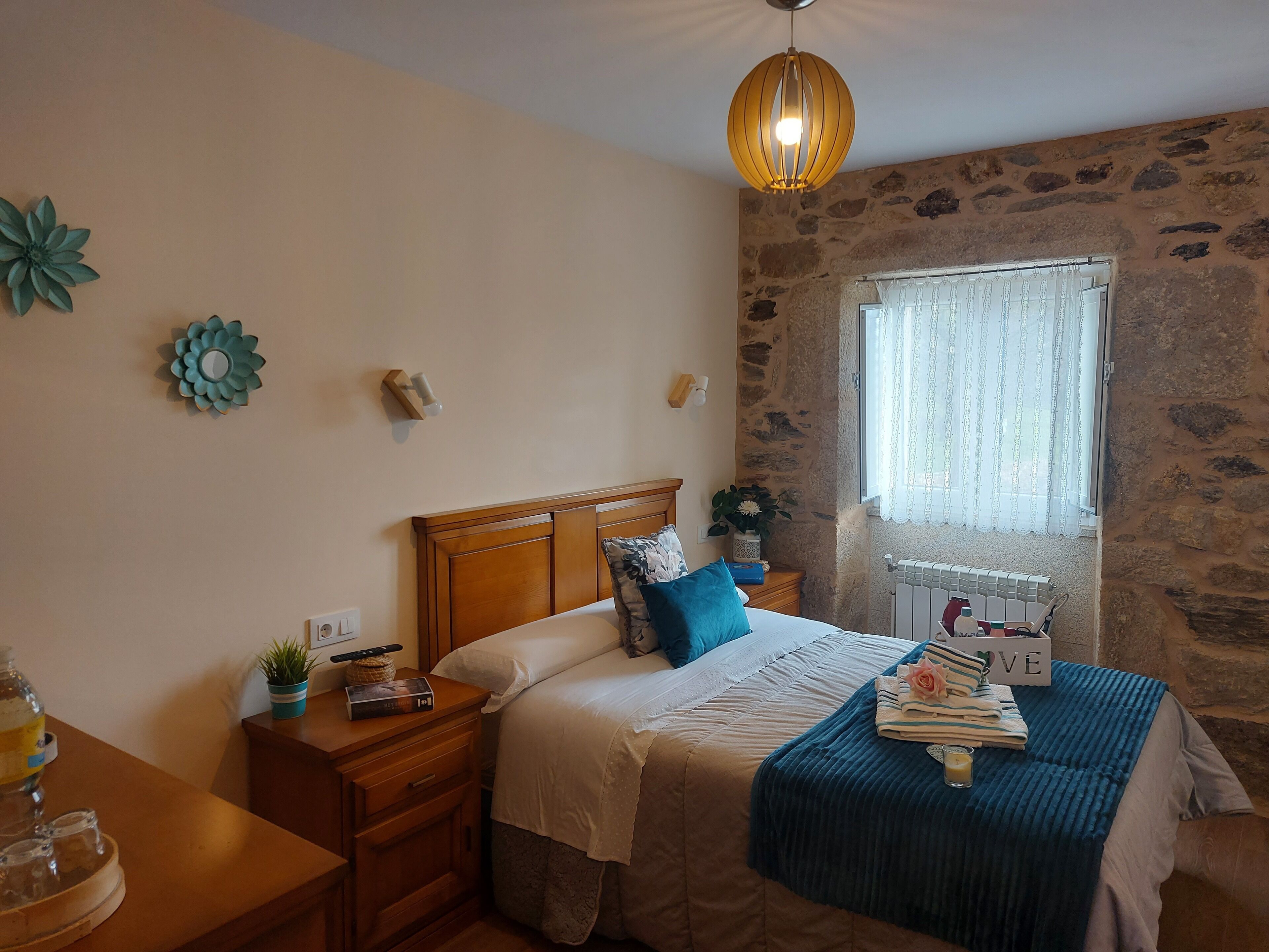 Quarto com parede de pedra, cama de madeira, mesa de cabeceira com luminária e janela com cortinas.