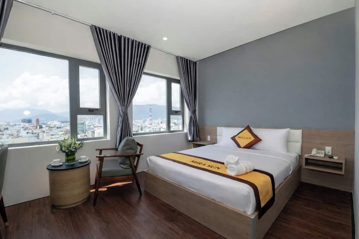 Quarto de hotel com uma cama grande, uma cadeira, uma mesinha com um vaso de flores e uma mesa de cabeceira com um telefone.