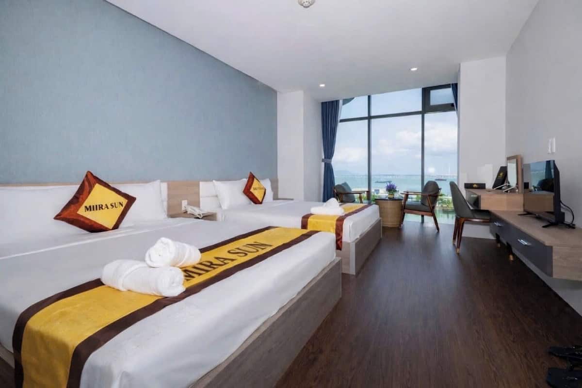 Quarto de hotel com duas camas, uma escrivaninha e vista para o mar.