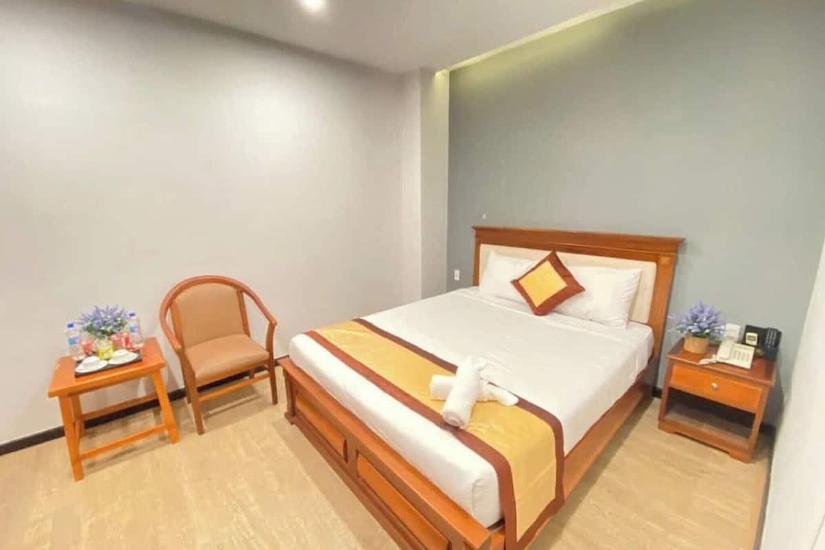 Quarto de hotel com cama de madeira, uma cadeira, uma mesinha com artigos de higiene pessoal e um criado-mudo com telefone e uma planta.