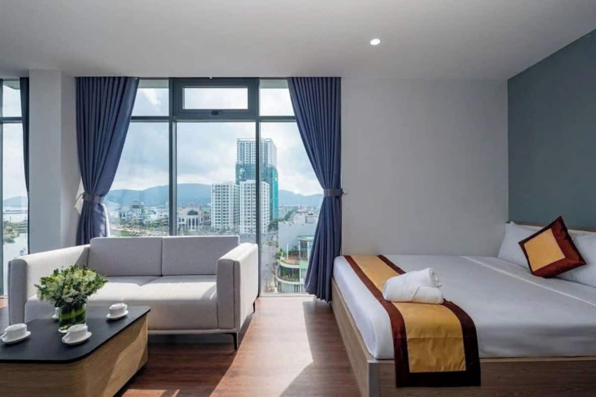 Quarto de hotel com cama, sofá, mesa de centro e vista para a paisagem urbana.