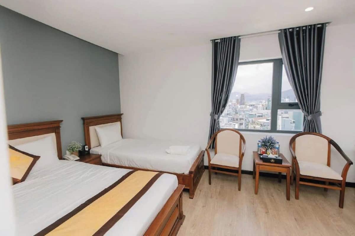 Quarto de hotel com duas camas, uma mesa pequena com um vaso e vista para a cidade através de amplas janelas.