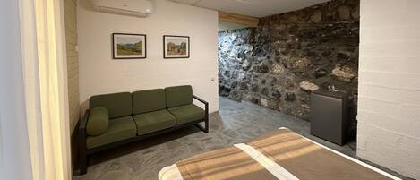 Um quarto com uma cama, um sofá verde, duas imagens emolduradas, uma parede de pedra e um aparelho de ar condicionado.