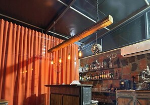 Uma área de bar com prateleiras de madeira repletas de garrafas, um lustre pendurado e uma janela grande com cortinas.