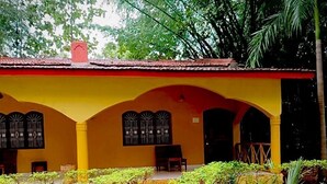 Um edifício com fachada amarela e telhado vermelho, rodeado por vegetação exuberante e um pequeno espelho de água.