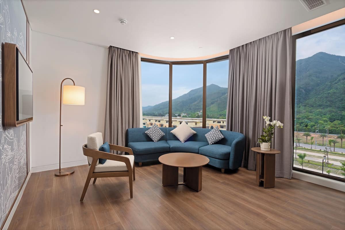 Quarto de hotel moderno com uma janela ampla que oferece vista para as montanhas, um sofá azul com almofadas estampadas, piso de madeira e uma pequena mesa com um vaso de planta.