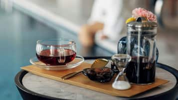 Uma mesa com um copo de bebida vermelha, um pote com uma flor e uma pequena tigela com comida, dispostos num hall de hotel com piscina ao fundo.