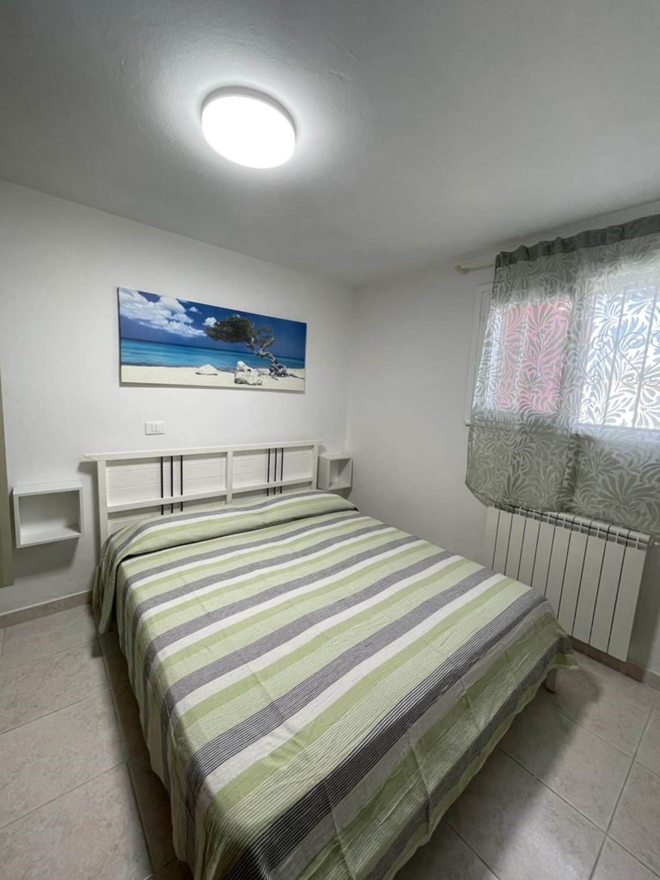 Quarto com cama, quadro de uma praia, janela com cortinas estampadas e radiador.