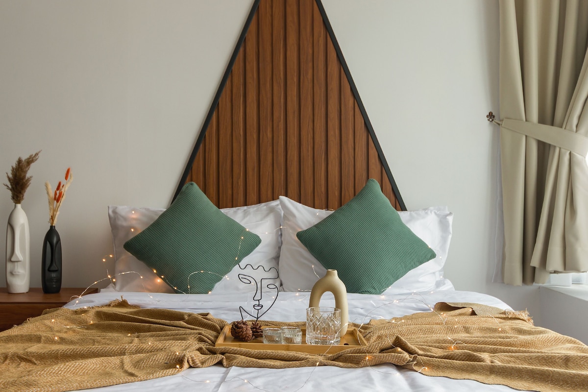 Cama bem arrumada com cabeceira de madeira, travesseiros verdes e um vaso decorativo com plantas secas em uma mesa de cabeceira.