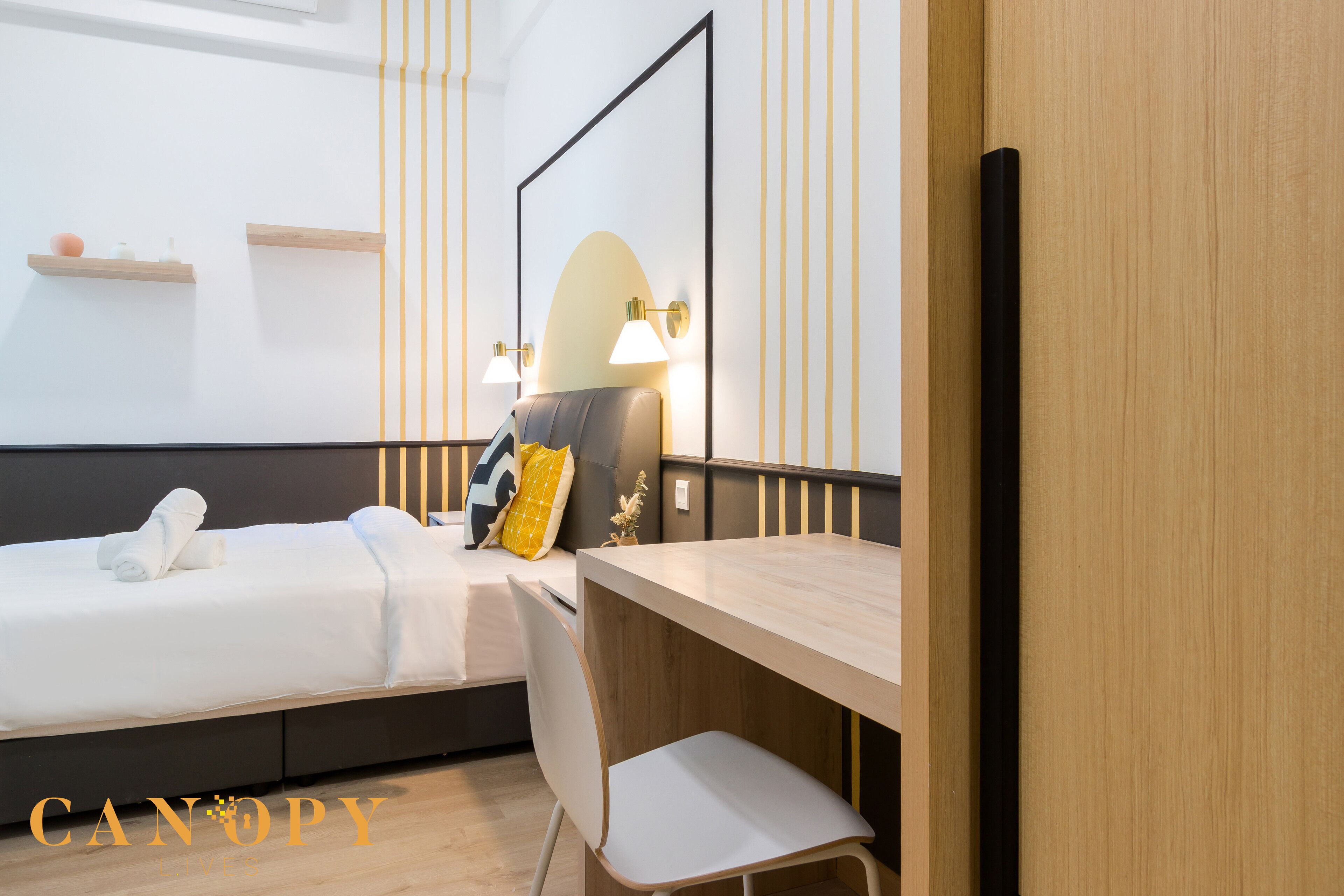Quarto de hotel com cama, escrivaninha, cadeira, espelho e prateleira com produtos de higiene pessoal.