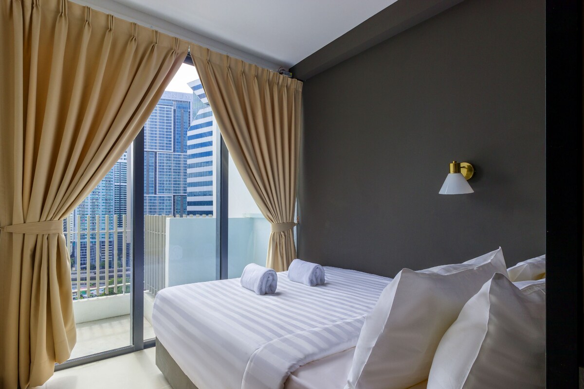 Quarto de hotel com uma cama grande, duas almofadas, uma mesa de cabeceira, uma luminária fixada na parede e vista para a paisagem urbana através de janelas do chão ao teto.