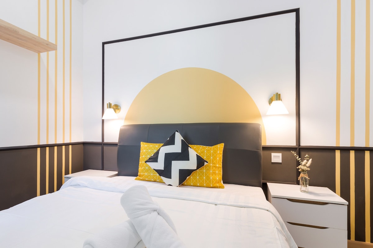Quarto de hotel com cama, mesa de cabeceira, abajur e parede com listras verticais.