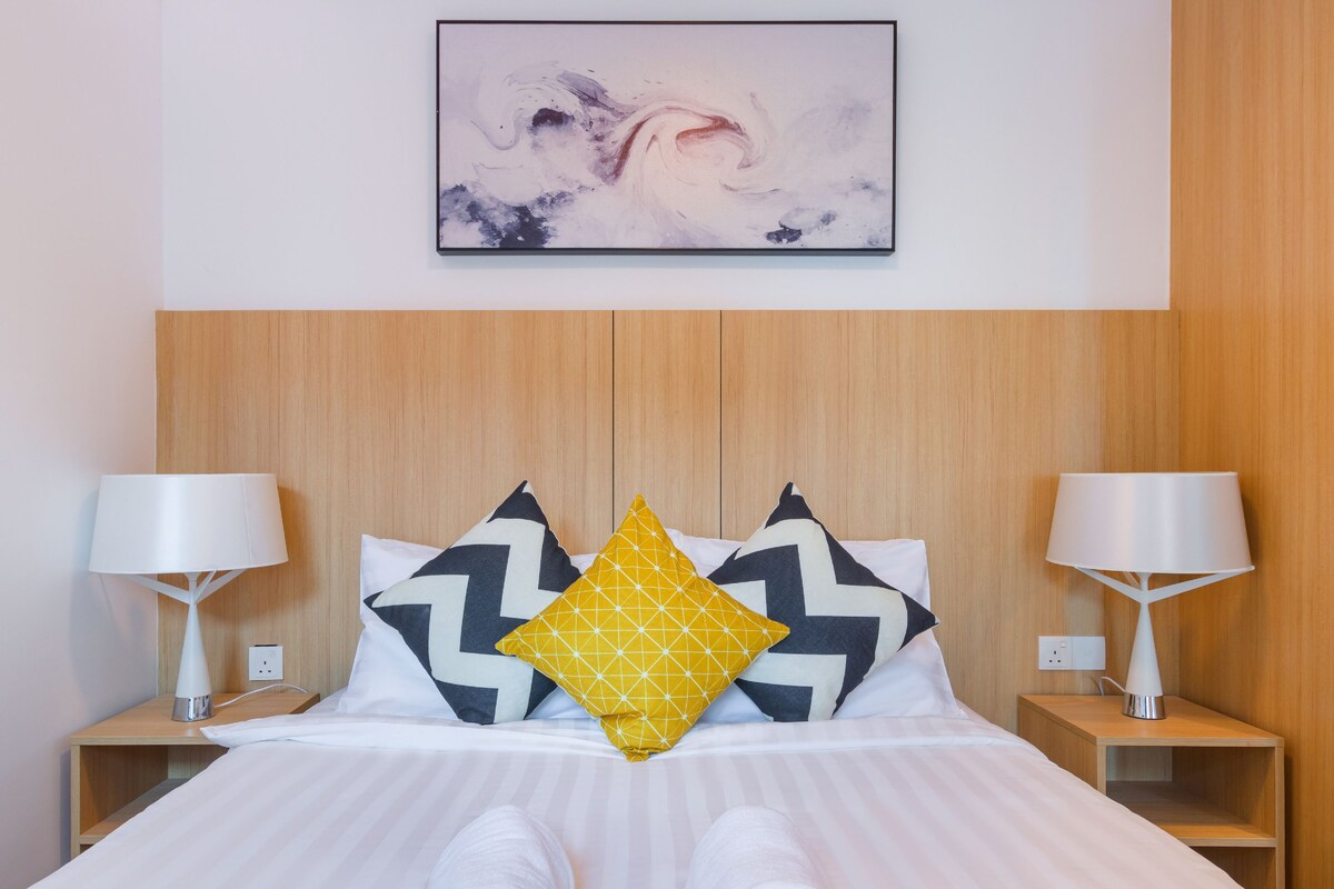 Quarto de hotel com uma cama, duas mesas de cabeceira com abajures, um quadro na parede e uma tomada visível.