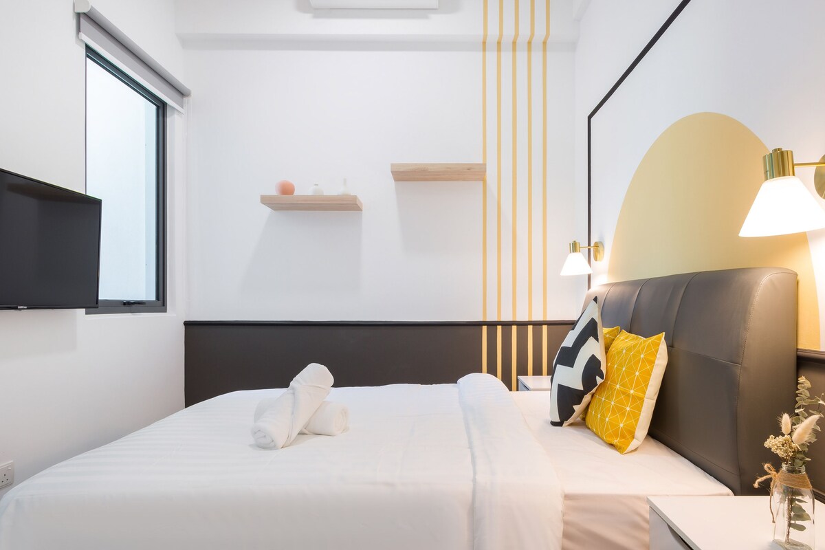 Uma cama bem arrumada com edredom branco e travesseiros, uma mesinha de cabeceira com um vaso, e um abajur fixado na parede.