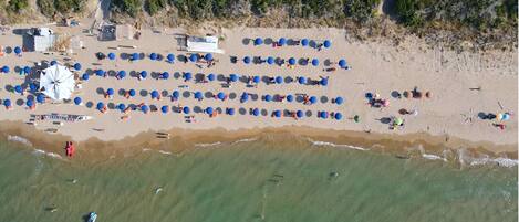 Vista aérea de uma praia com fileiras de guarda-sóis azuis e cadeiras de praia, uma torre de salva-vidas e alguns barcos perto da costa.