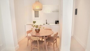 Uma área de refeições com uma mesa redonda, quatro cadeiras de madeira, um lustre pendurado e uma cozinha ao fundo.