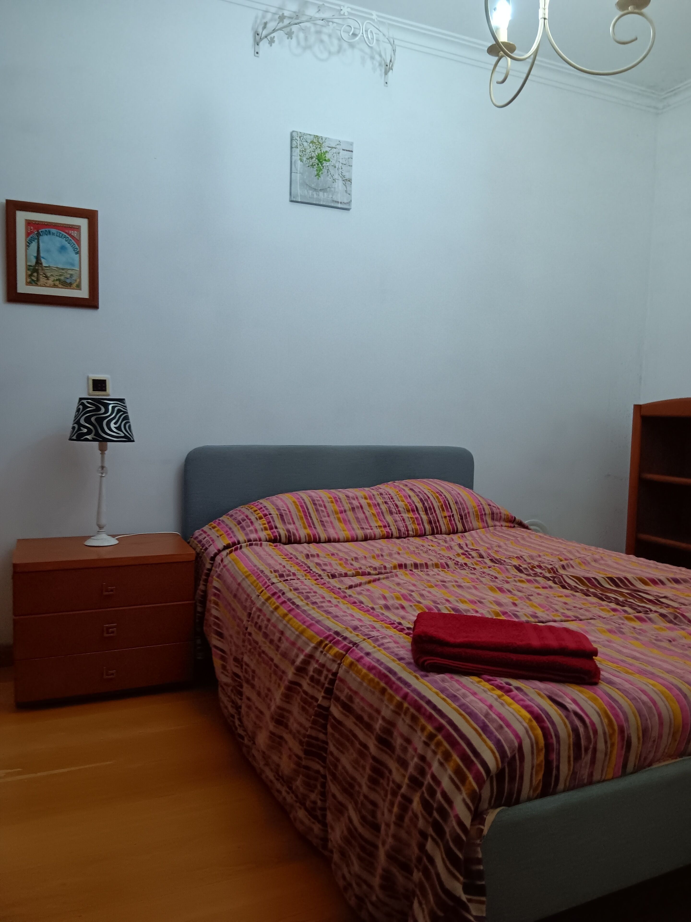 Quarto com cama, mesa de cabeceira, luminária, porta-retrato e lustre.