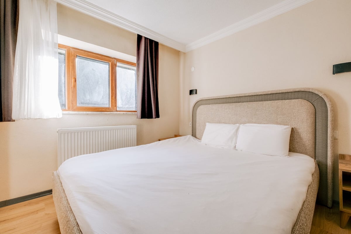 Cama bem arrumada em quarto com janela, radiador e luminária fixada na parede.